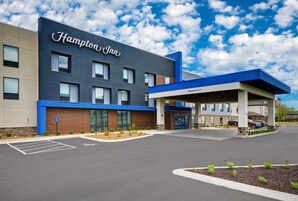 Um hotel Hampton Inn com fachada moderna, área de estacionamento coberta e céu claro.