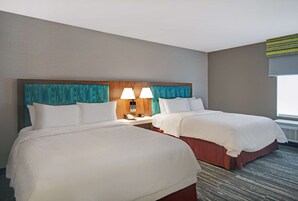 Quarto de hotel com duas camas, cabeceira de madeira, mesa de cabeceira com luminárias e janela com vista.