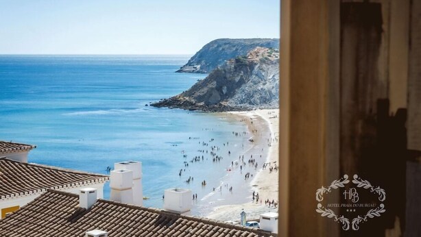 Vista costeira com praia, falésias e edifícios com telhados em terracota.