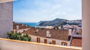 Uma vila costeira com edifícios de telhados em terracota, uma montanha ao fundo e vista para o mar.