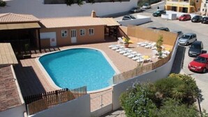 Piscina com espreguiçadeiras, área cercada e estacionamento com carros.