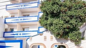 Hotel com fachada branca e detalhes em azul, apresentando uma placa grande e uma árvore em frente.