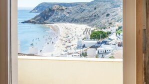 Vista costeira de uma janela com vista para uma praia, edifícios brancos e uma paisagem acidentada.