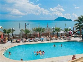 Uma área de piscina com pessoas nadando e relaxando, cercada por palmeiras e bandeiras, com vista para o mar azul e límpido.