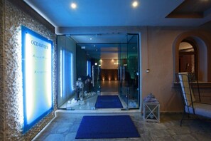 Um saguão de hotel com uma mesa de recepção, entrada por porta de vidro e um tapete azul que leva à área da recepção.