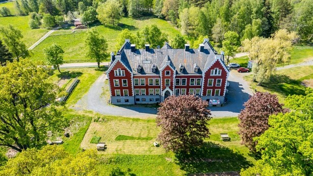 Um edifício grande, com vários andares e fachada simétrica, cercado por vegetação exuberante e um gramado bem cuidado.