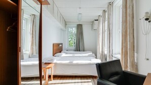 Quarto compacto com uma cama de solteiro, uma mesinha, uma cadeira e uma janela com cortinas.