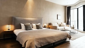 Quarto de hotel moderno com uma cama grande, um sofá menor, uma mesa de cabeceira e uma janela ampla com cortinas.