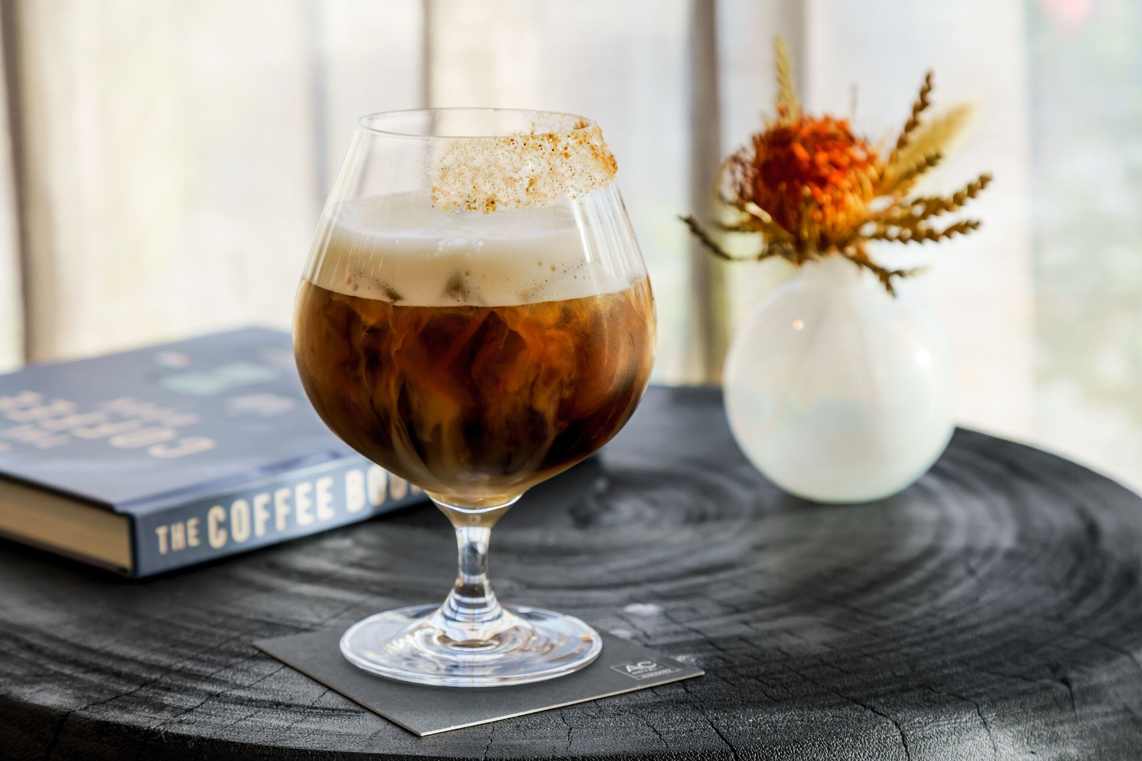 Um copo de café gelado sobre uma mesa de madeira, com um livro intitulado 