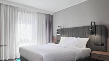 Quarto de hotel moderno com uma cama grande, mesa de cabeceira, luminária e vista para o exterior através de cortinas translúcidas.