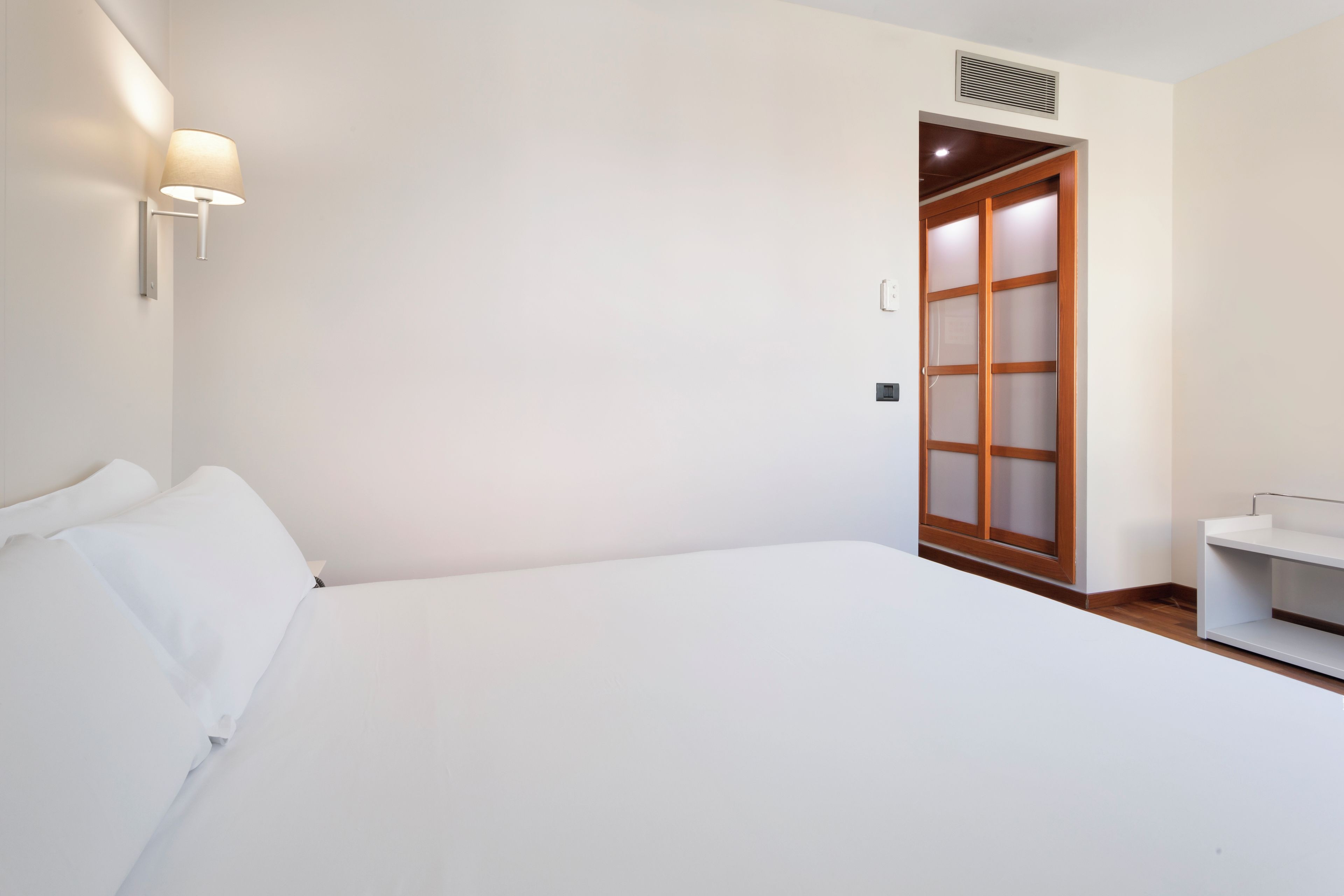 Quarto com uma cama grande, uma mesa de cabeceira, uma luminária na parede e uma porta de correr com painéis de vidro jateado.