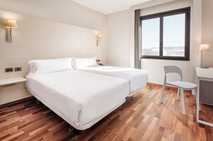 Quarto de hotel com duas camas, uma escrivaninha, uma cadeira e uma janela grande.