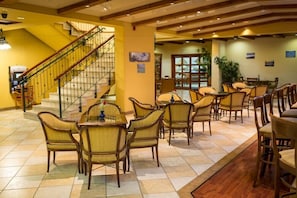 Um saguão de hotel com uma escadaria, diversas mesas de jantar com cadeiras e piso ladrilhado.