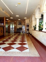 Um hall de hotel com piso xadrez, balcão de recepção, escada e estante de livros.