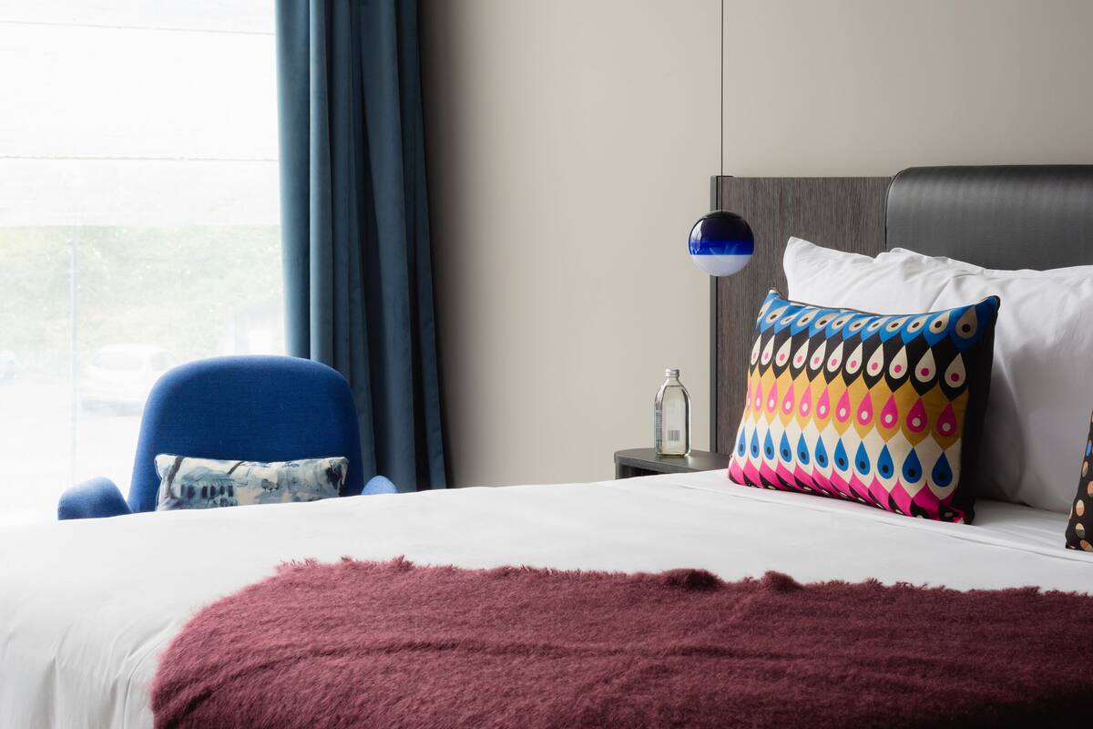 Quarto de hotel moderno com uma cama grande, uma cadeira azul, mesa de cabeceira com abajur e vista para o exterior.