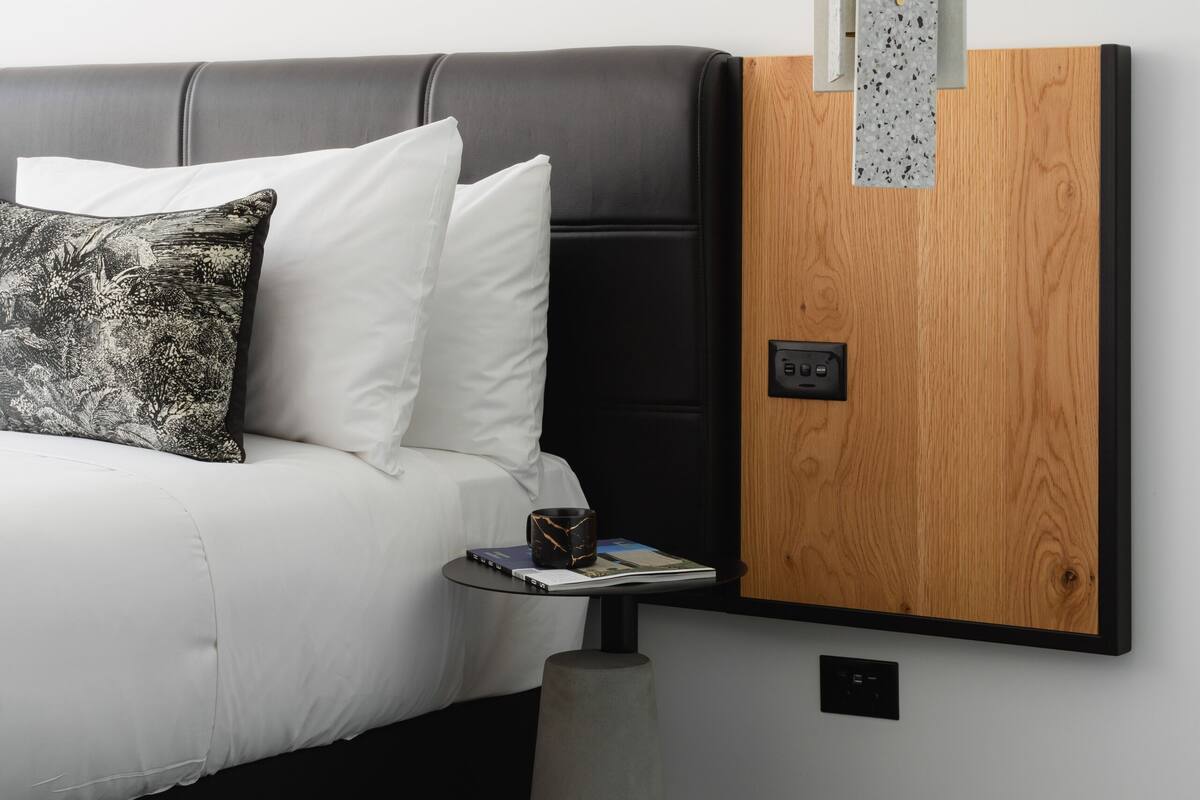 Quarto moderno com cama, mesa de cabeceira com abajur e painel de madeira na parede.