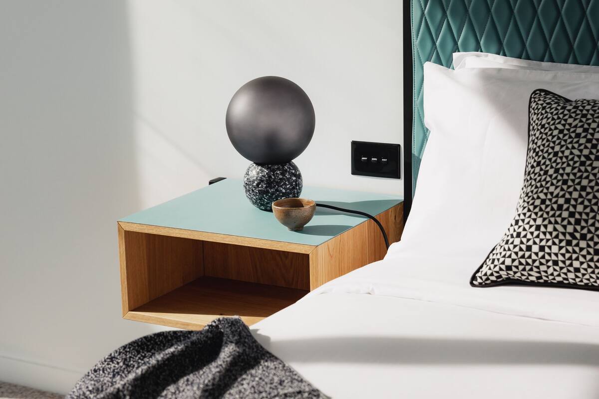 Uma cama bem arrumada com cabeceira verde-água, uma mesa de cabeceira com um objeto esférico e uma tigela, e um travesseiro estampado.