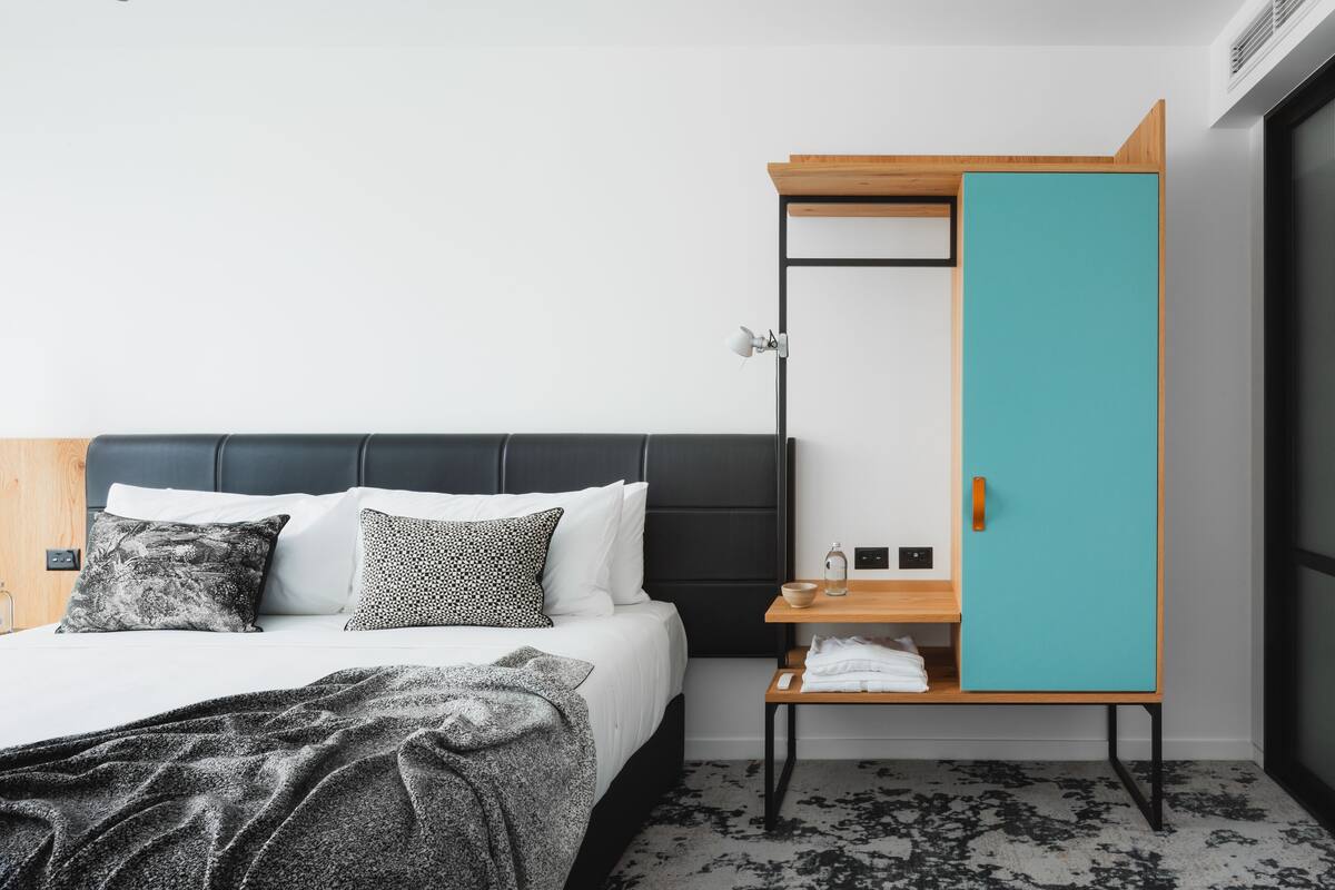 Quarto de hotel moderno com cama, guarda-roupa, mesa de cabeceira e abajur.