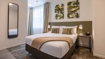 Quarto de hotel com uma cama grande, mesa de cabeceira com abajur, espelho e arte abstrata na parede.