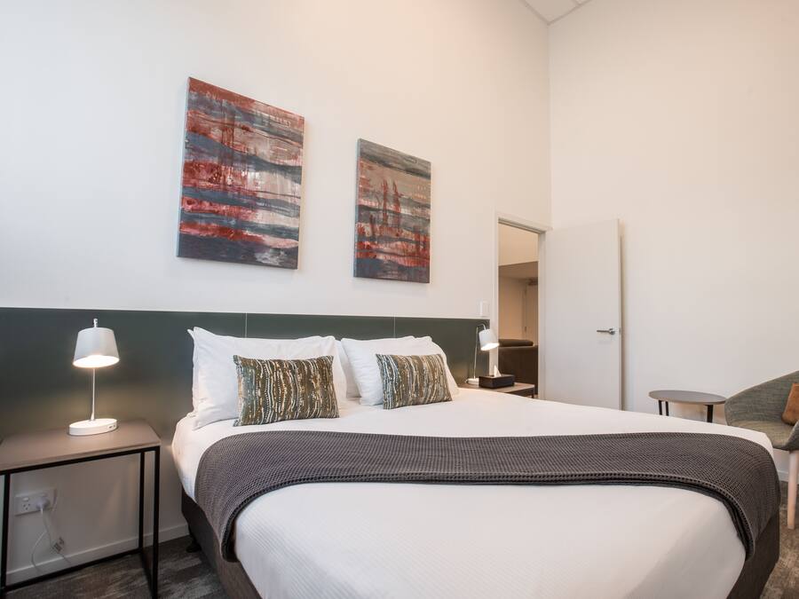 Quarto de hotel com uma cama grande, duas mesas de cabeceira com abajures, uma cadeira e duas pinturas abstratas na parede.