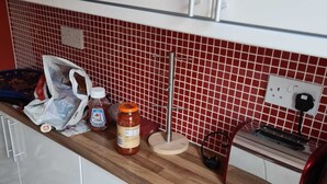 Uma cozinha com armários brancos, bancada de madeira e revestimento de azulejos vermelhos. Há potes com condimentos, uma chaleira e um prato com desenho floral.