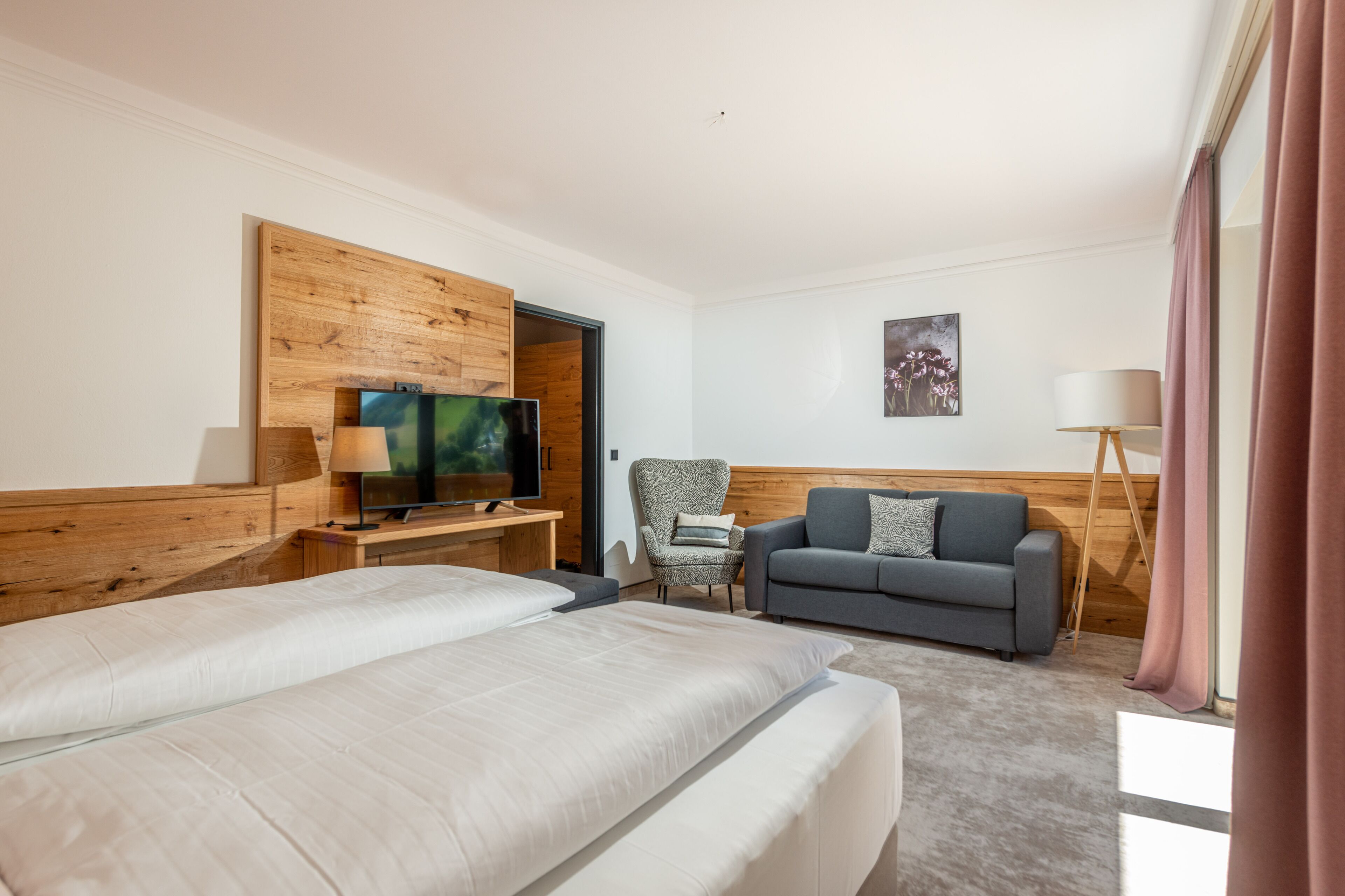 Quarto de hotel com duas camas, televisão, sofá e uma parede de destaque em madeira.