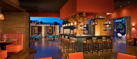 Uma área de bar moderna, com balcão, banquetas e um espaço de estar com mesas e cadeiras.