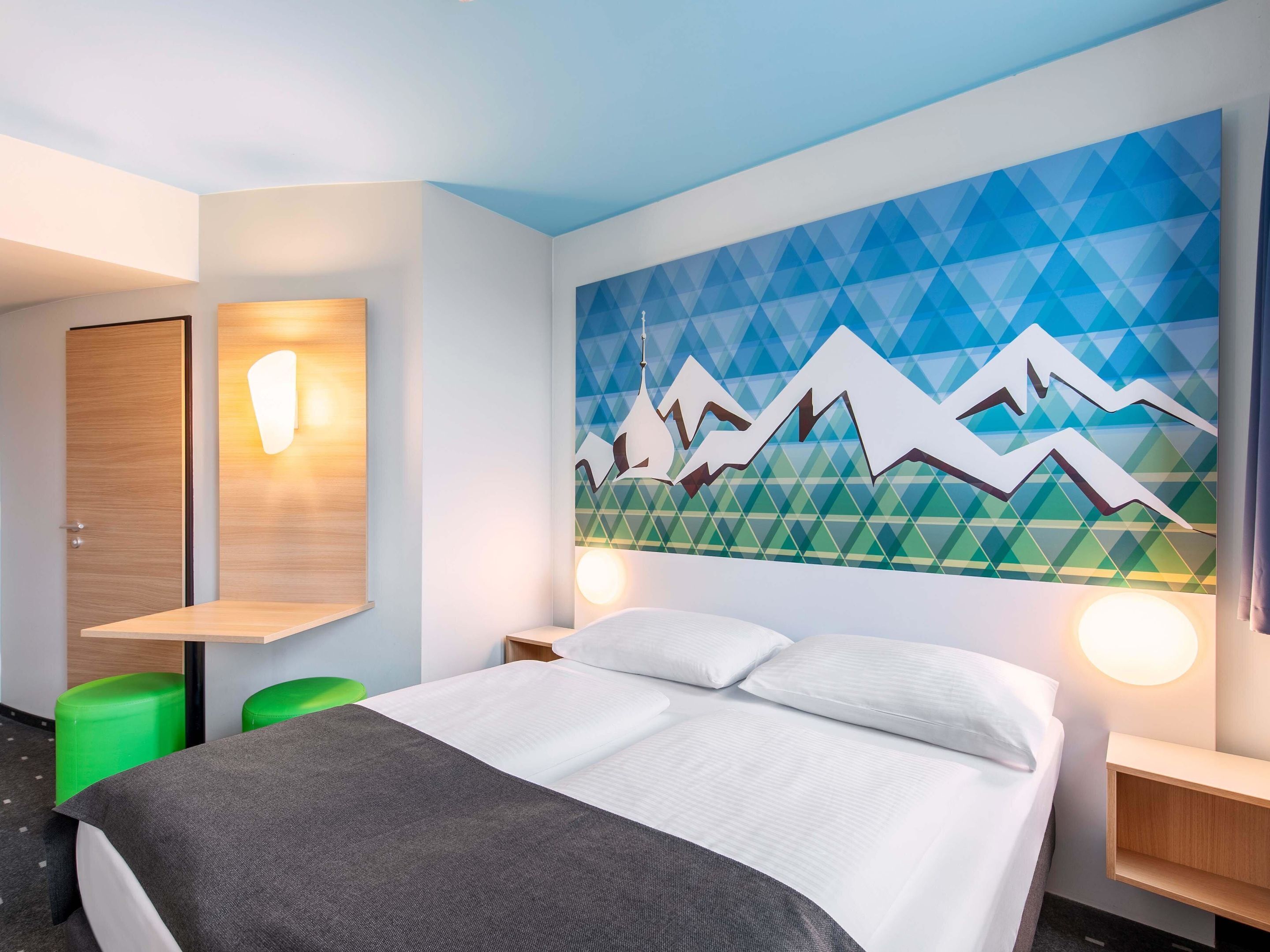 Quarto de hotel com uma cama, um banquinho verde, uma mesa de cabeceira de madeira e uma parede com um desenho geométrico de montanhas.