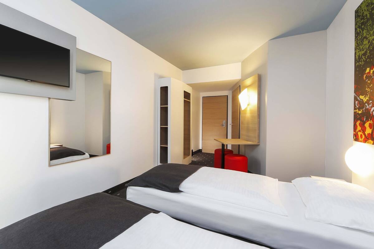 Quarto de hotel moderno com cama, televisão presa na parede, espelho e um banquinho vermelho.