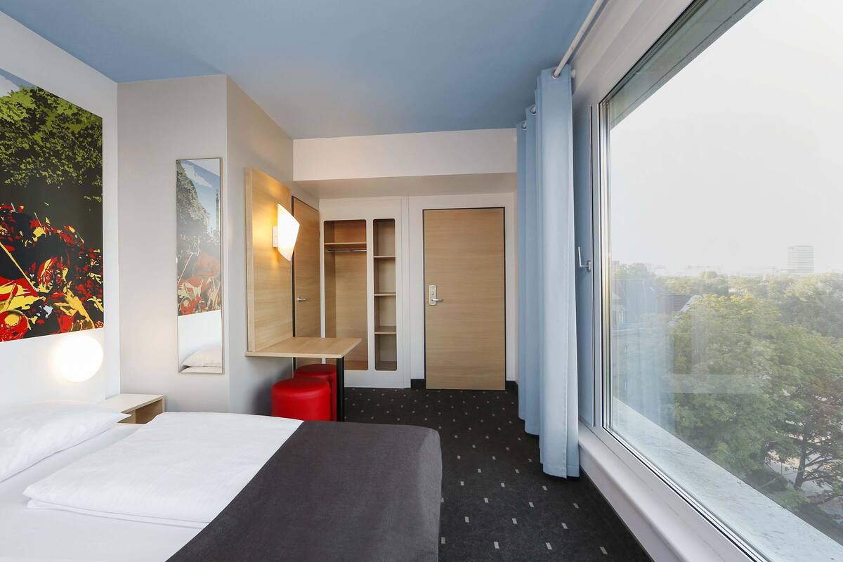 Quarto de hotel moderno com cama, escrivaninha, guarda-roupa e uma janela grande com vista para a cidade.
