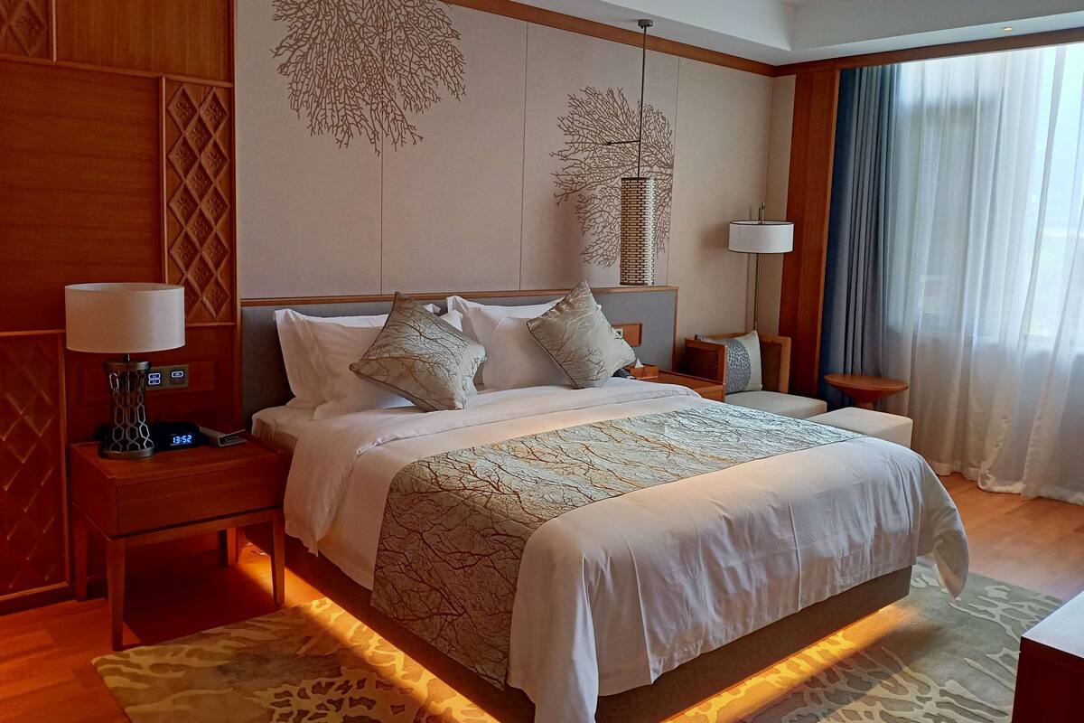 Quarto de hotel com uma cama grande, mesa de cabeceira com abajur, obra de arte na parede e janela com cortinas.