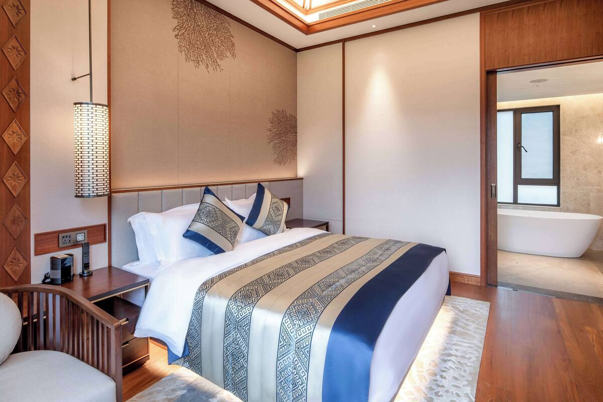 Quarto de hotel moderno com uma cama grande, uma mesa de cabeceira com abajur, uma mesinha de madeira e vista para um banheiro com banheira.