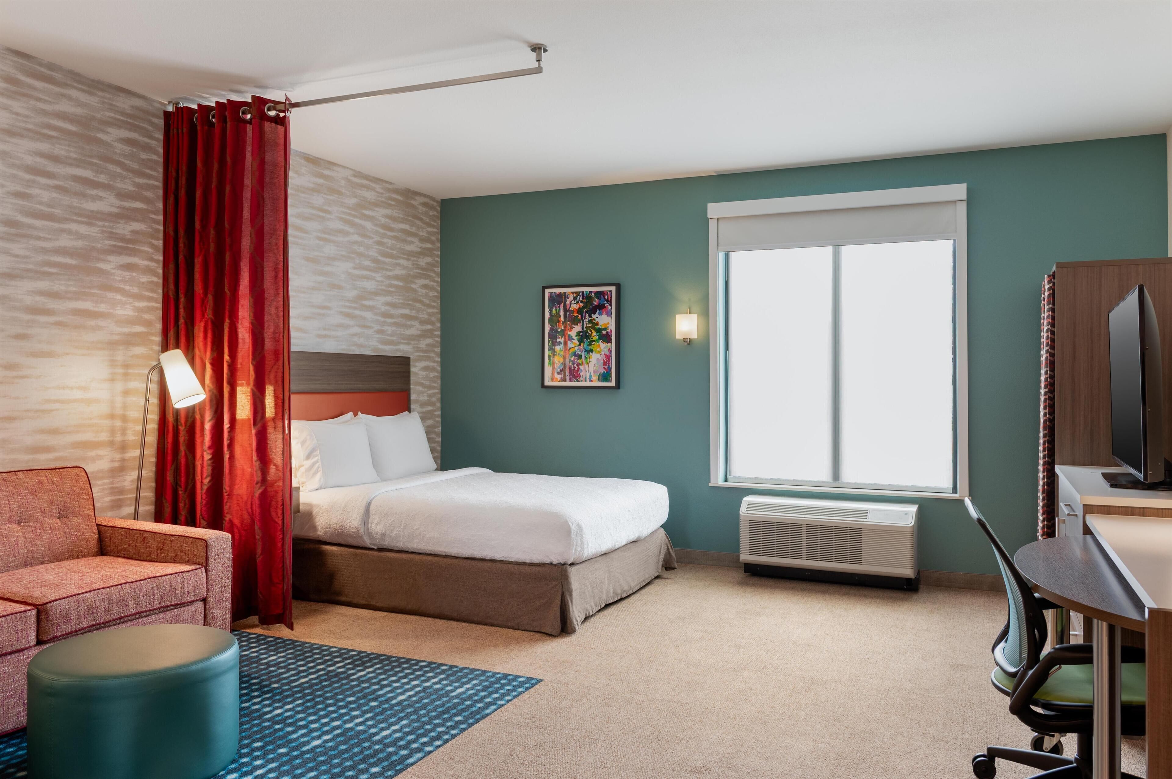 Quarto de hotel com cama, escrivaninha, cadeira, luminária, janela e um quadro na parede.