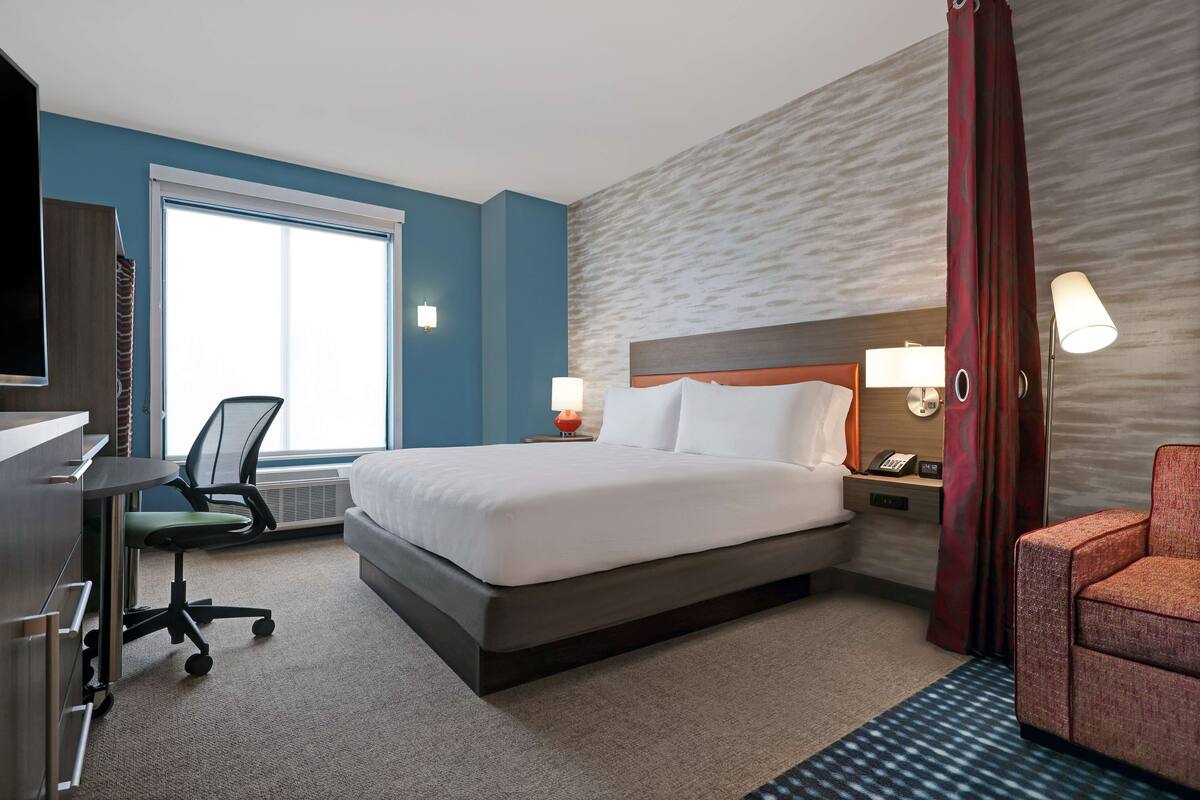 Quarto de hotel com cama, escrivaninha, cadeira e uma janela grande.