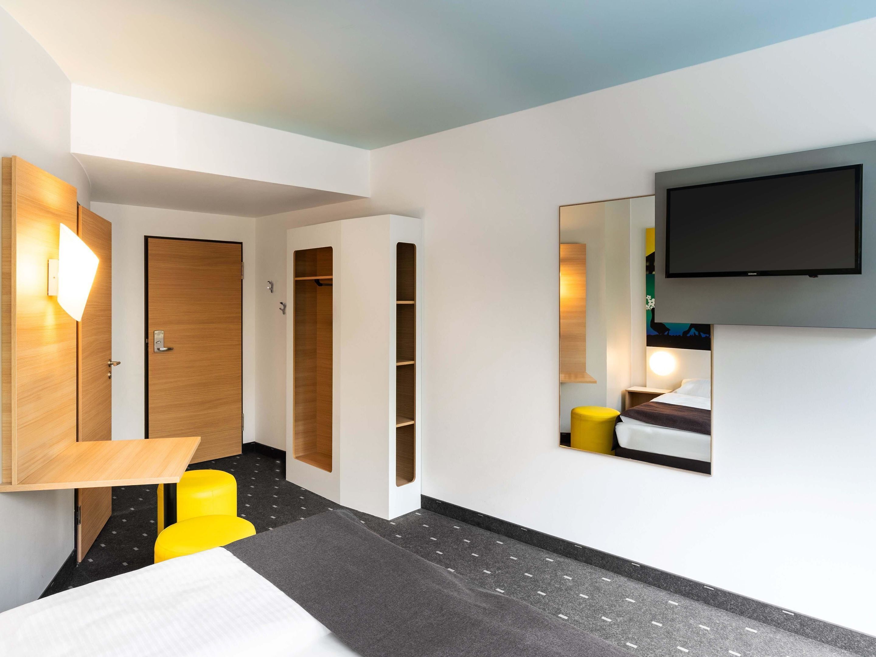 Quarto de hotel moderno com cama, escrivaninha, guarda-roupa, espelho e televisão.