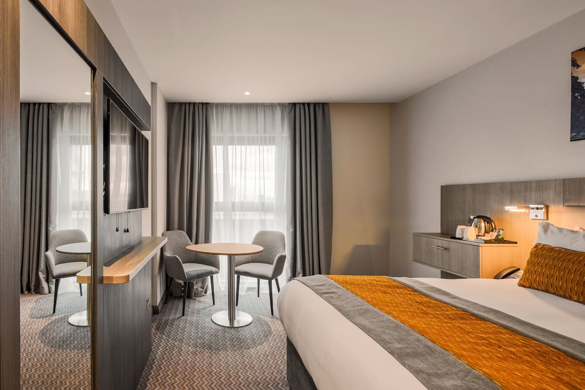 Quarto de hotel com cama, escrivaninha, duas cadeiras, televisão e mesa de cabeceira.