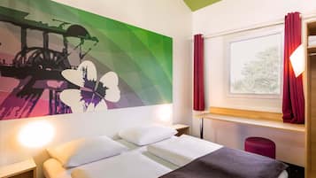 Quarto de hotel com cama, mesa de cabeceira, janela com cortinas e parede com um mural colorido.