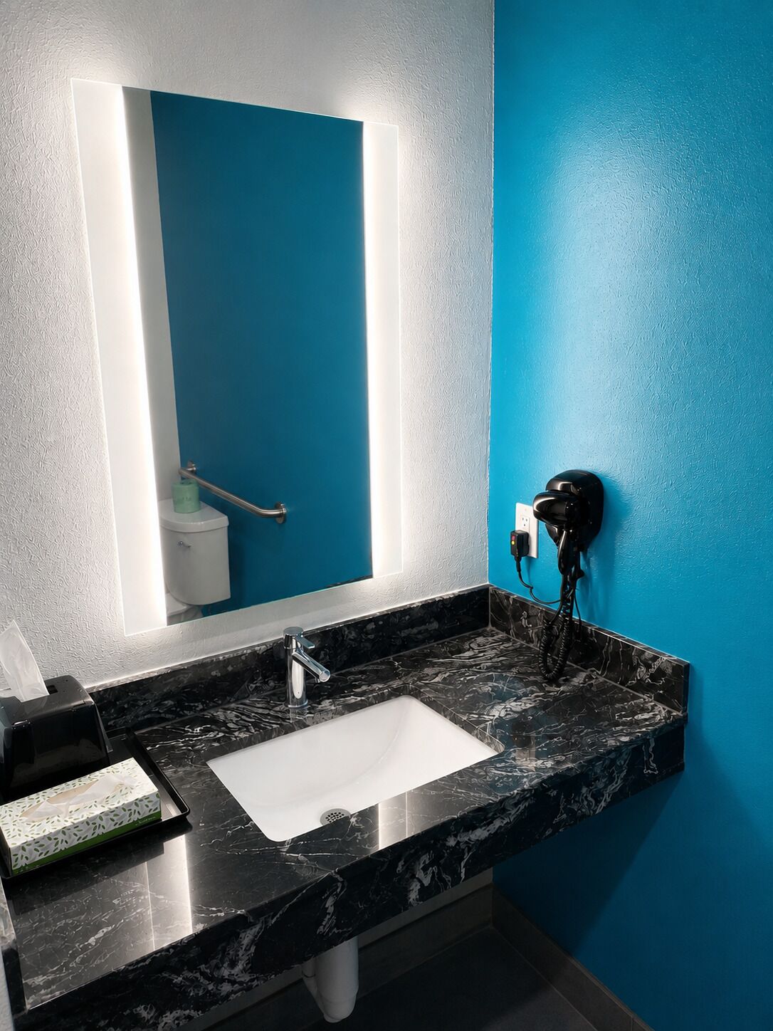 Um banheiro com pia branca, bancada de mármore preto, parede azul e espelho com iluminação.
