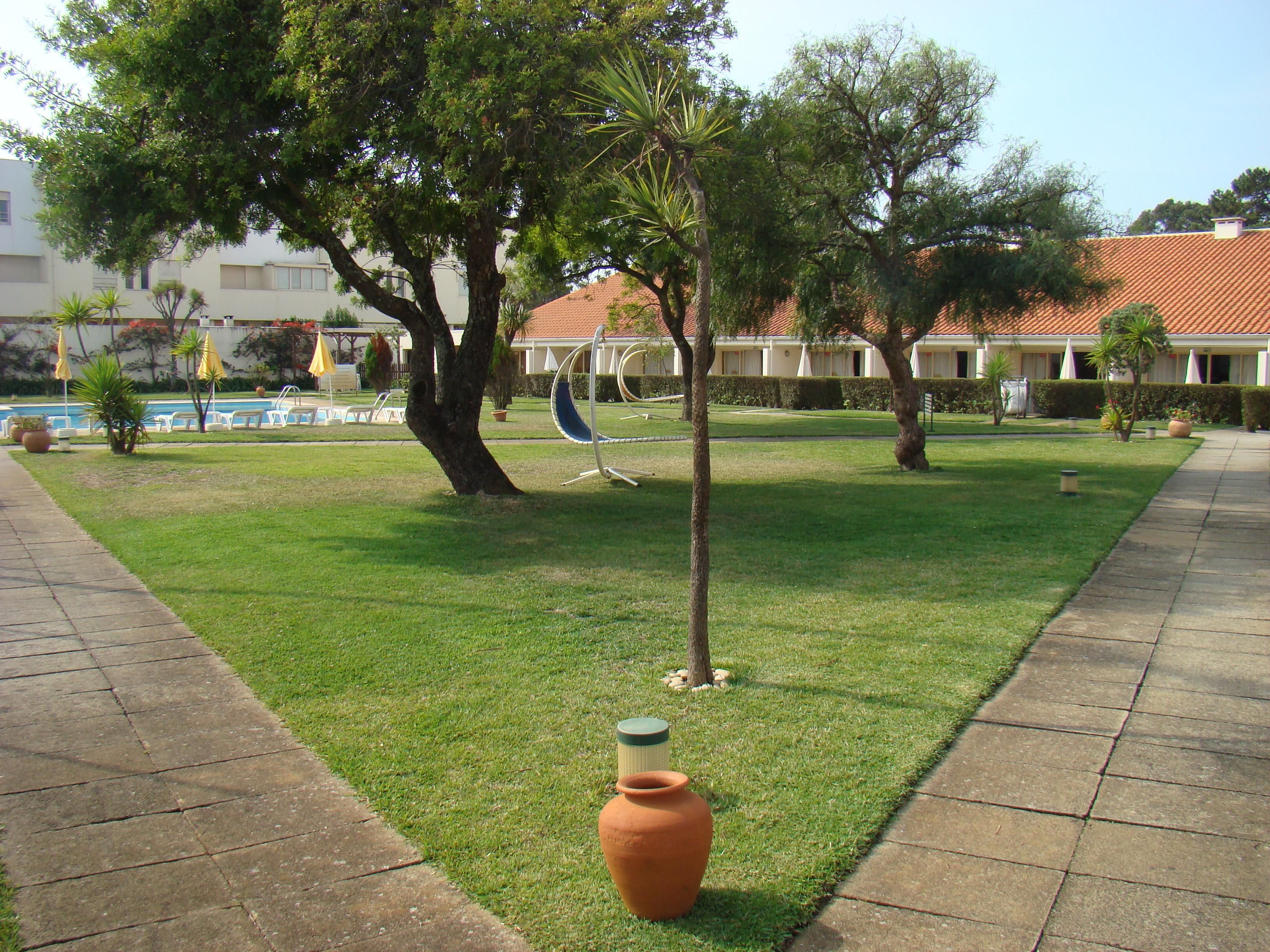 Uma área gramada bem cuidada com uma grande árvore, um balanço e um vaso decorativo.