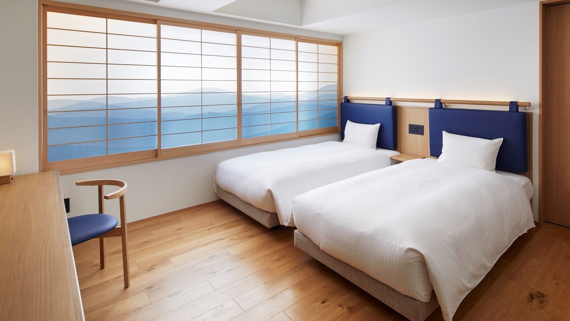 Quarto de hotel com duas camas, uma escrivaninha, uma cadeira e uma janela grande com vista para montanhas.