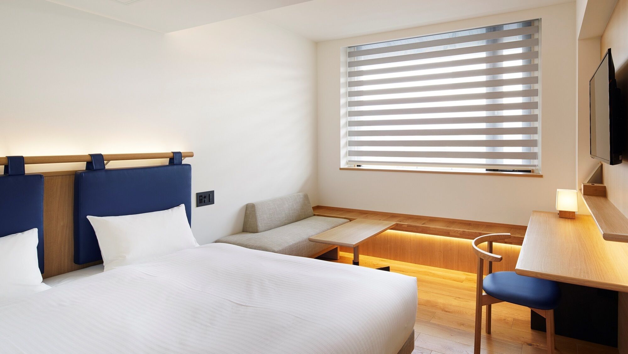 Quarto de hotel com cama, escrivaninha, cadeira, sofá e janela com persianas.