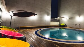 Piscina interior com piso de madeira, dois guarda-sóis e um banco.