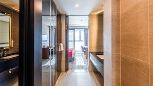Um corredor moderno de hotel com uma porta de vidro que dá para um quarto com cama, escrivaninha e cadeira.