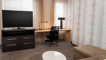 Quarto de hotel com televisão de tela plana, uma escrivaninha com cadeira, um sofá e uma mesa redonda pequena.