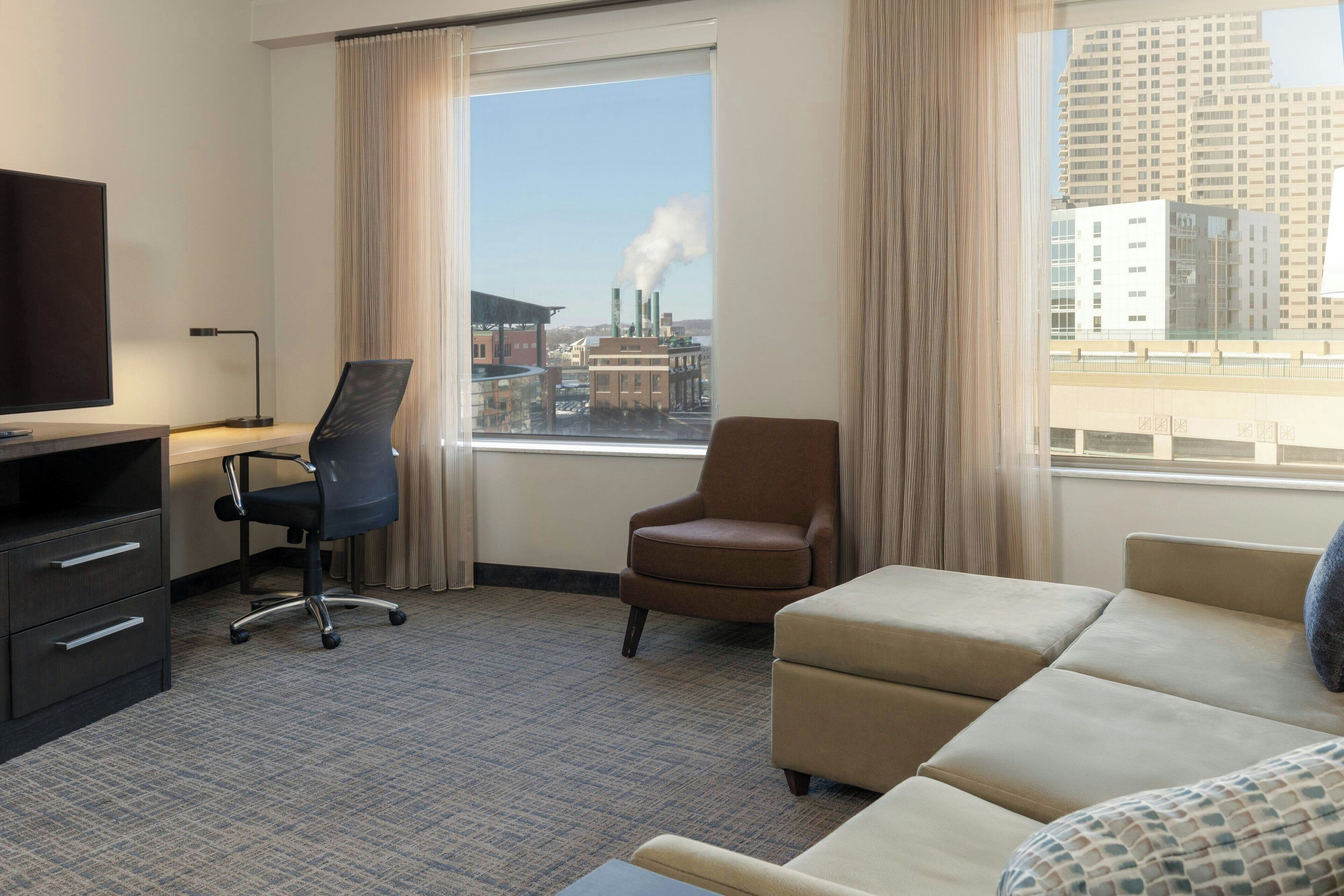 Quarto de hotel com uma janela grande, uma escrivaninha com cadeira, um sofá e uma televisão.