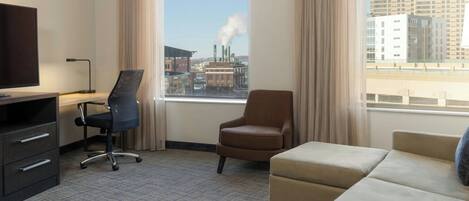 Quarto de hotel com uma janela grande, uma escrivaninha com cadeira, um sofá e uma televisão.