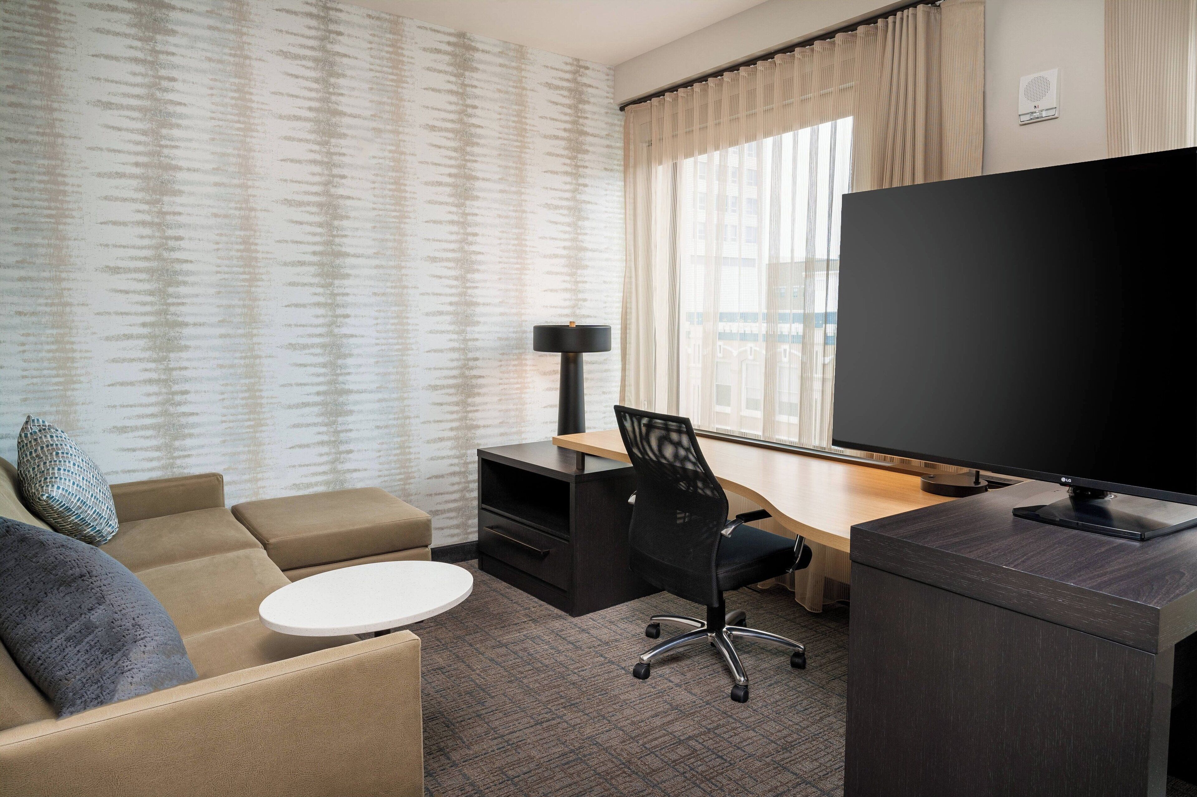 Quarto de hotel com sofá, escrivaninha com cadeira, televisão e janela com cortinas.