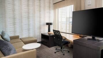 Quarto de hotel com sofá, escrivaninha com cadeira, televisão e janela com cortinas.