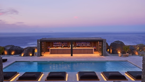 Bar à beira da piscina com vista para o oceano, rodeado por espreguiçadeiras e pedras grandes.
