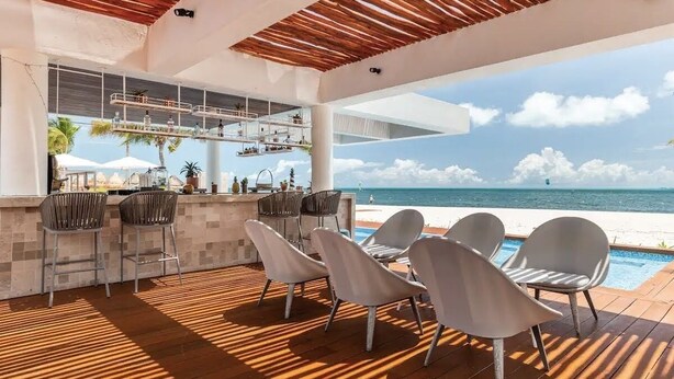 Área de bar externa com piscina e vista para a praia, apresentando assentos modernos e teto de madeira.